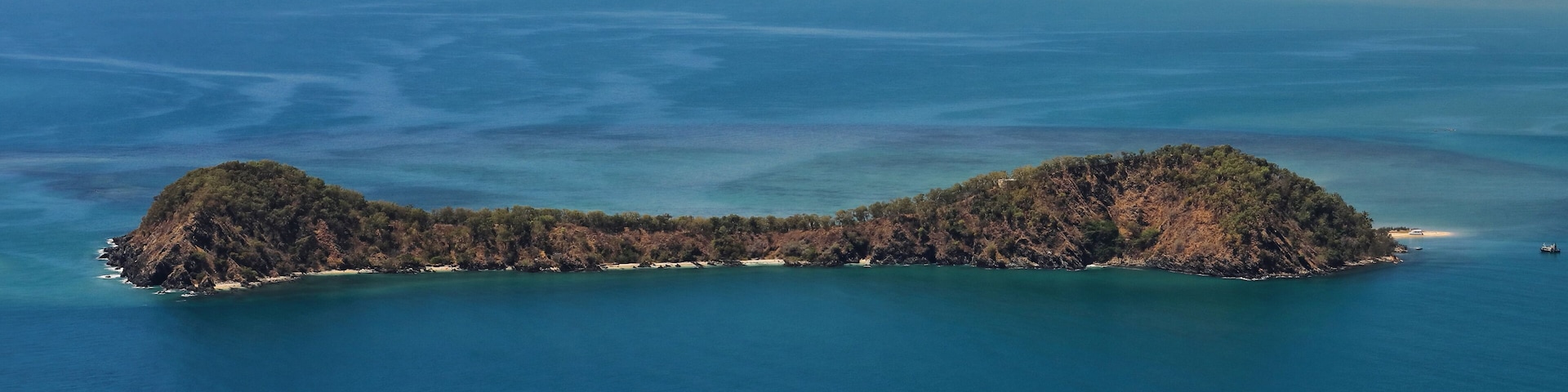 Vista aérea de uma ilha grande cercada por águas azuis cristalinas, com montanhas ao fundo.