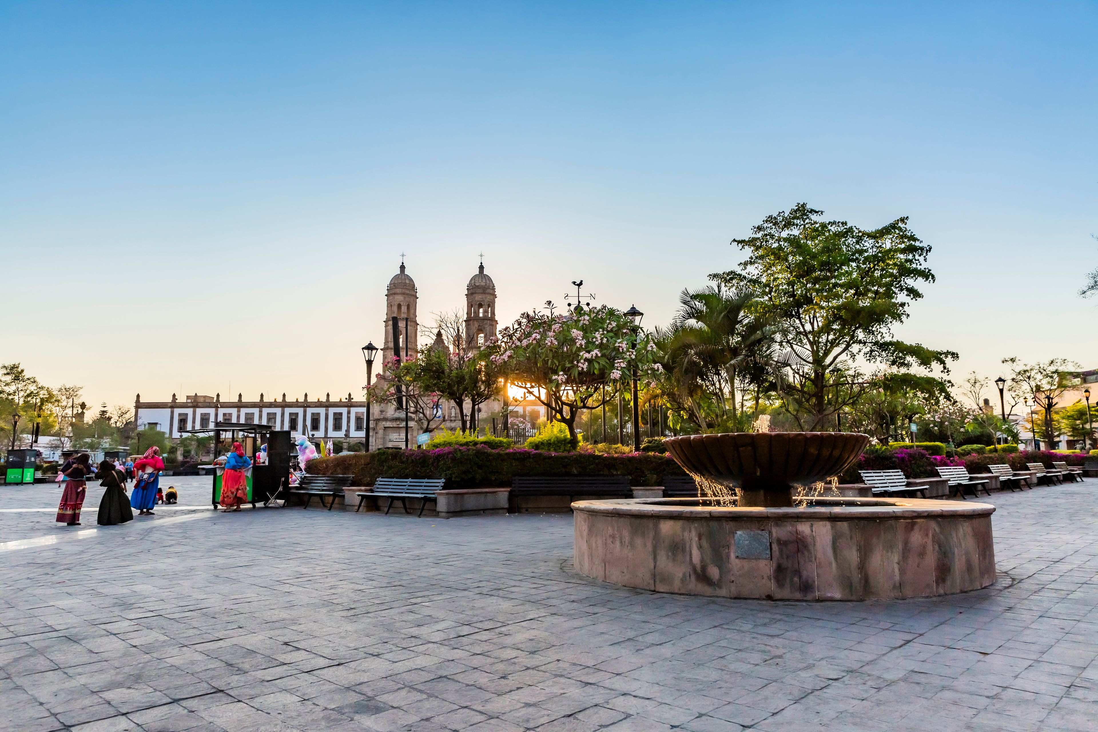 Uma praça com uma fonte, bancos e um edifício histórico com torres.
