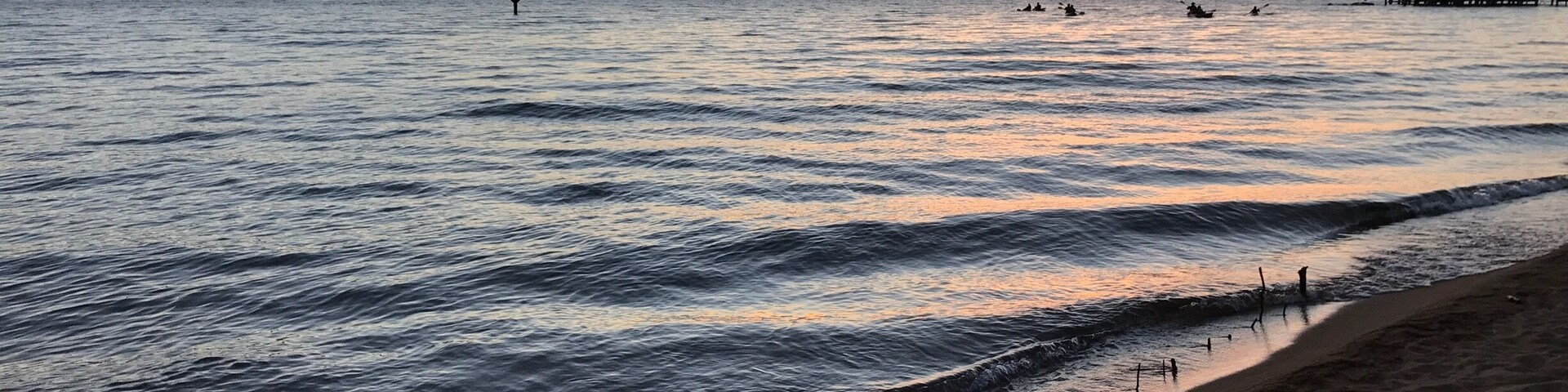 Uma praia ao pôr do sol, com águas calmas, areal arenoso e montanhas ao fundo.