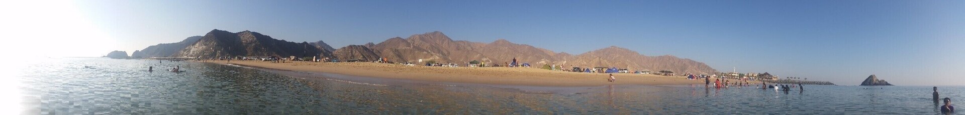 Uma praia com montanhas ao fundo, tendas e pessoas.