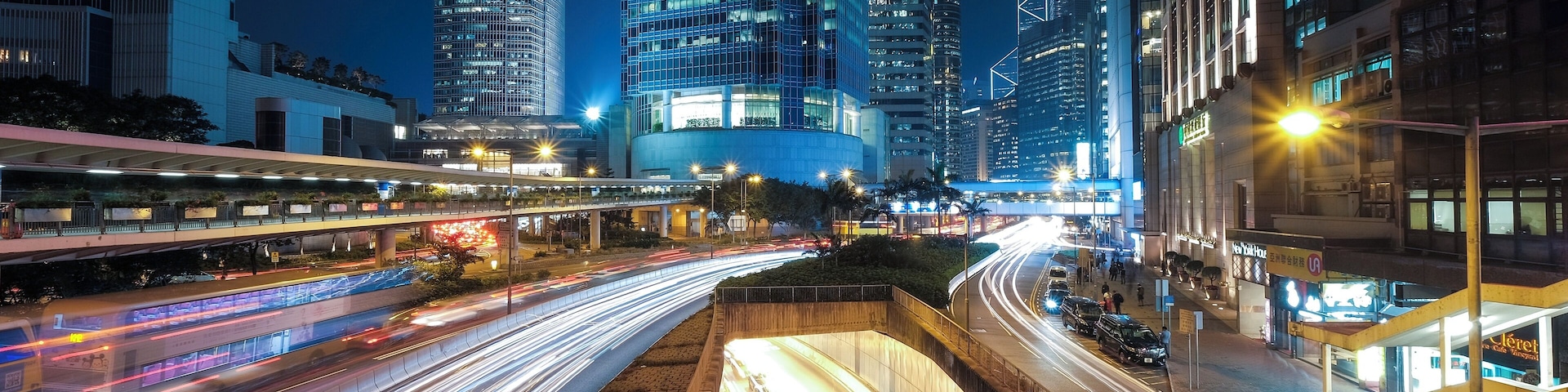 Paisagem urbana noturna com arranha-céus iluminados, uma movimentada autoestrada com múltiplas faixas e uma passarela suspensa para pedestres.