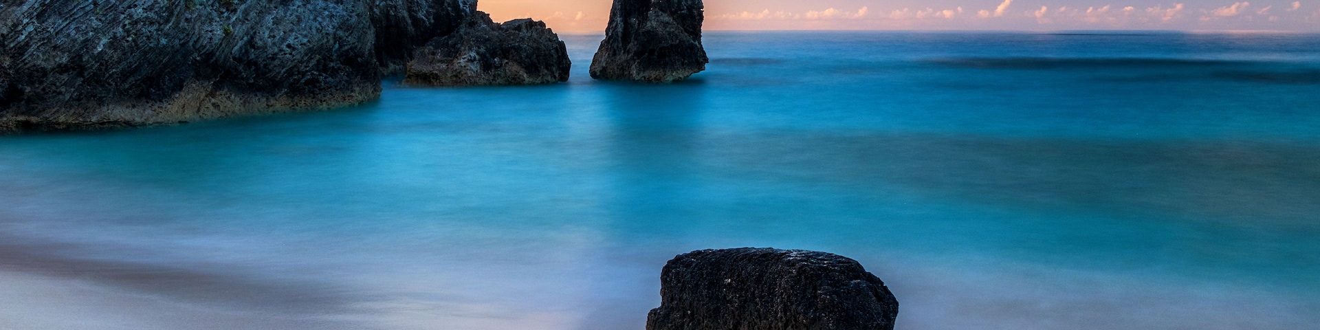 Paisagem costeira com uma formação rochosa imponente no mar, uma praia arenosa e um céu claro.