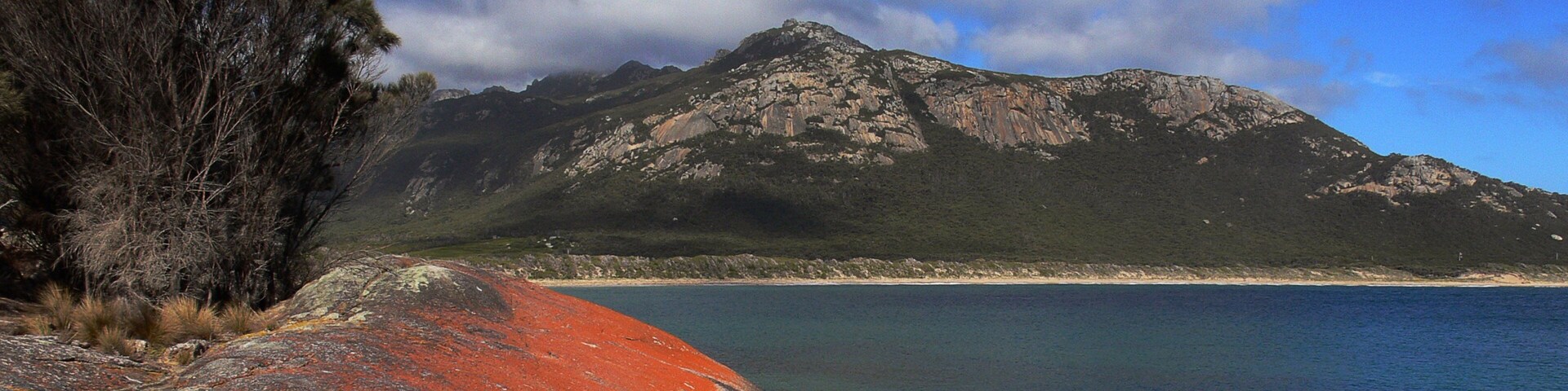 Paisagem costeira com um litoral rochoso, mar azul cristalino e uma montanha ao fundo.