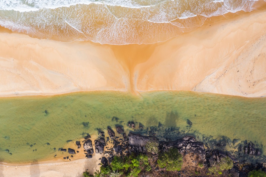 Uma praia com águas cristalinas, areal arenoso e rochas visíveis.