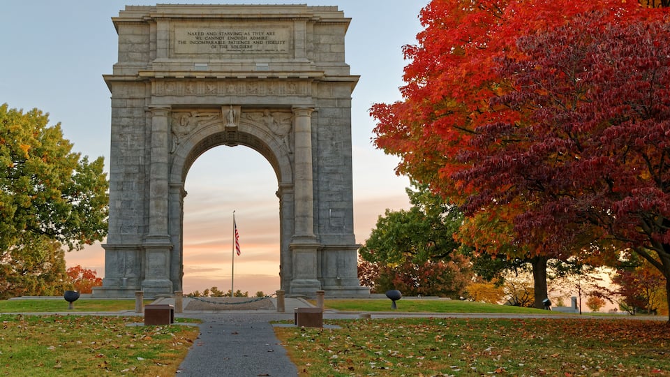 Monumento de pedra em arco com inscrições, cercado por folhagem outonal e uma bandeira americana.