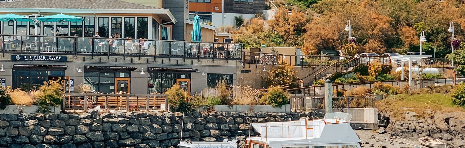 Marina com barcos atracados, um café à beira-mar e edifícios ao fundo.
