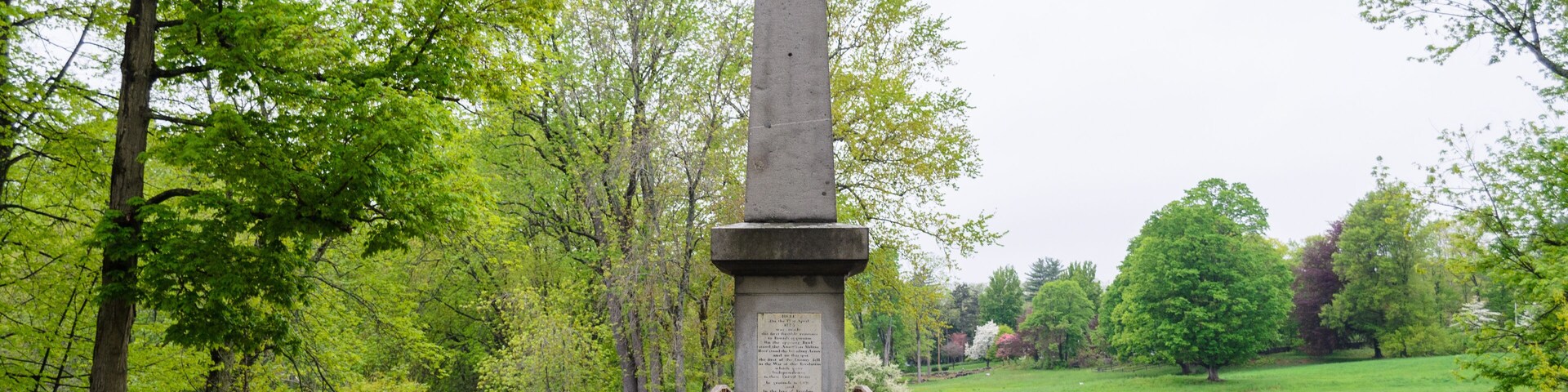 Um monumento alto e esguio com uma placa em um parque com árvores e um lago.