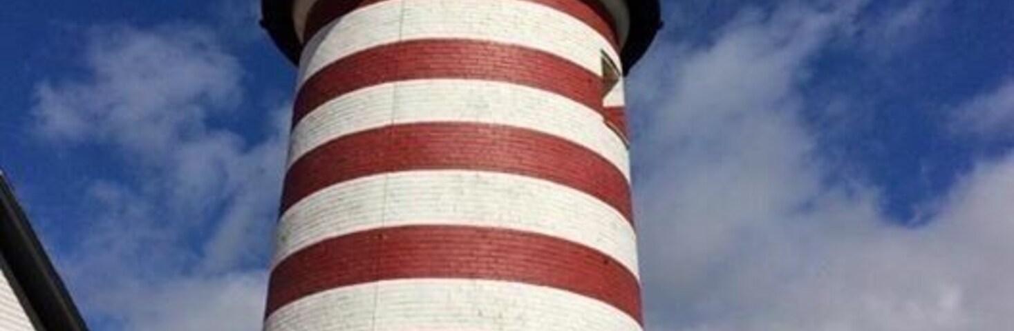 Um farol com listras vermelhas e brancas, um pequeno edifício branco ao lado e uma floresta ao fundo.