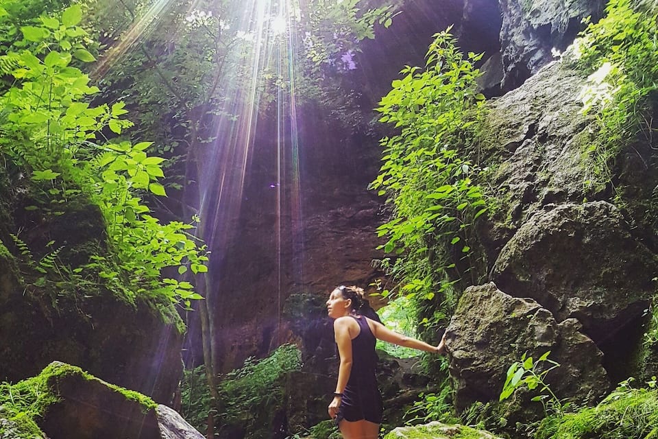 Uma pessoa em pé sobre uma rocha coberta de musgo em uma área arborizada, com a luz do sol filtrando pelas árvores.