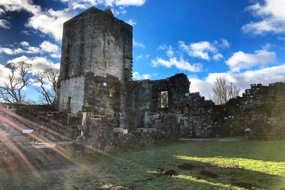 Uma torre de pedra antiga com uma bandeira no topo, rodeada por uma área gramada e árvores.
