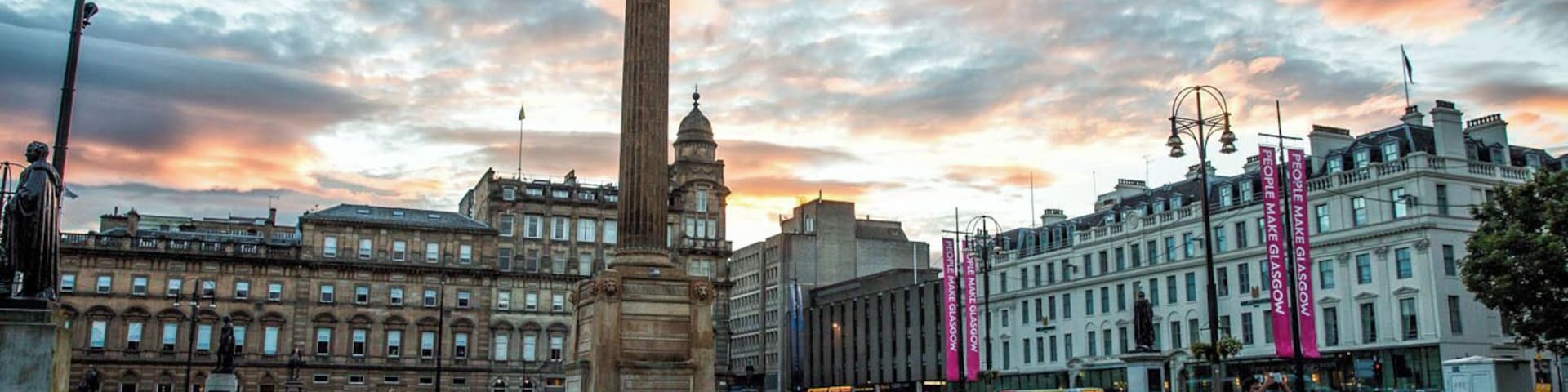 Uma praça urbana com um monumento imponente, edifícios históricos e um céu ao pôr do sol.
