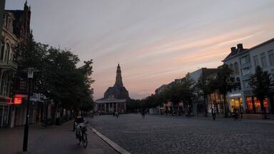 Uma rua de paralelepípedos com edifícios históricos, uma torre de igreja imponente e um céu ao pôr do sol.