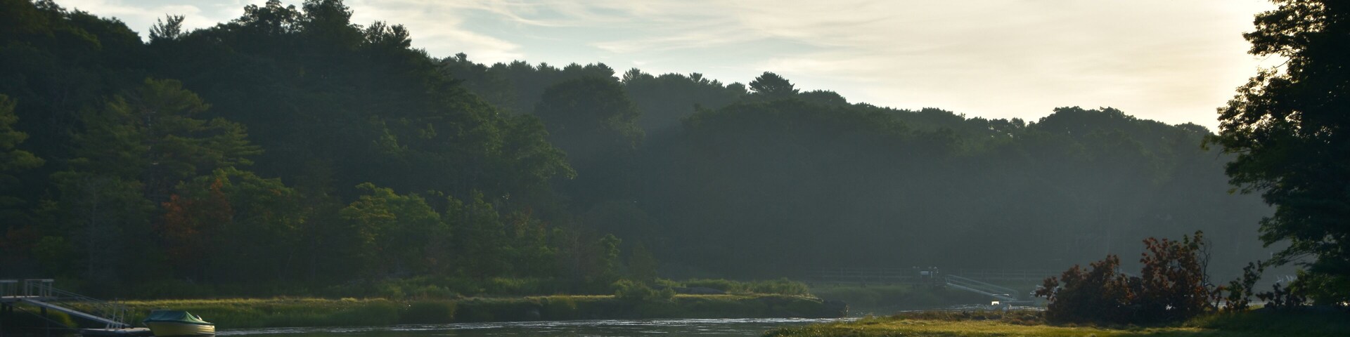 Um rio calmo com um pequeno barco, rodeado por uma floresta densa e uma colina nevoenta ao fundo.