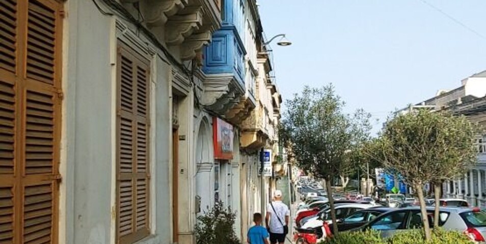 Uma rua de paralelepípedos estreita, ladeada por edifícios históricos, carros estacionados e vasos de plantas.