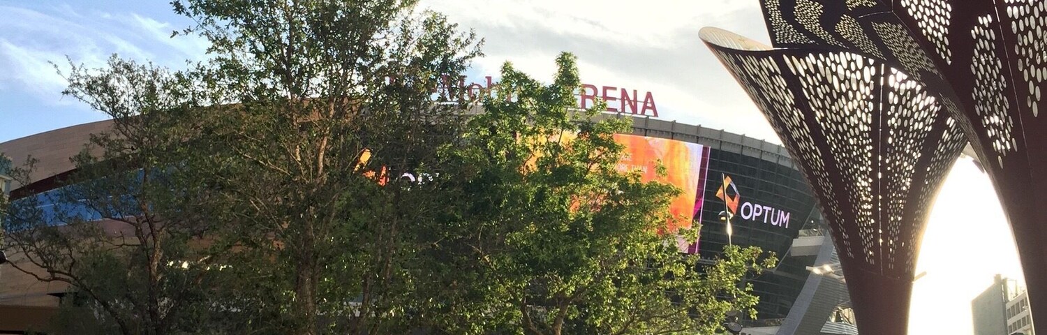 Uma praça pública com árvores, uma escultura moderna e um edifício com as placas 'BENA' e 'OPTUM'.