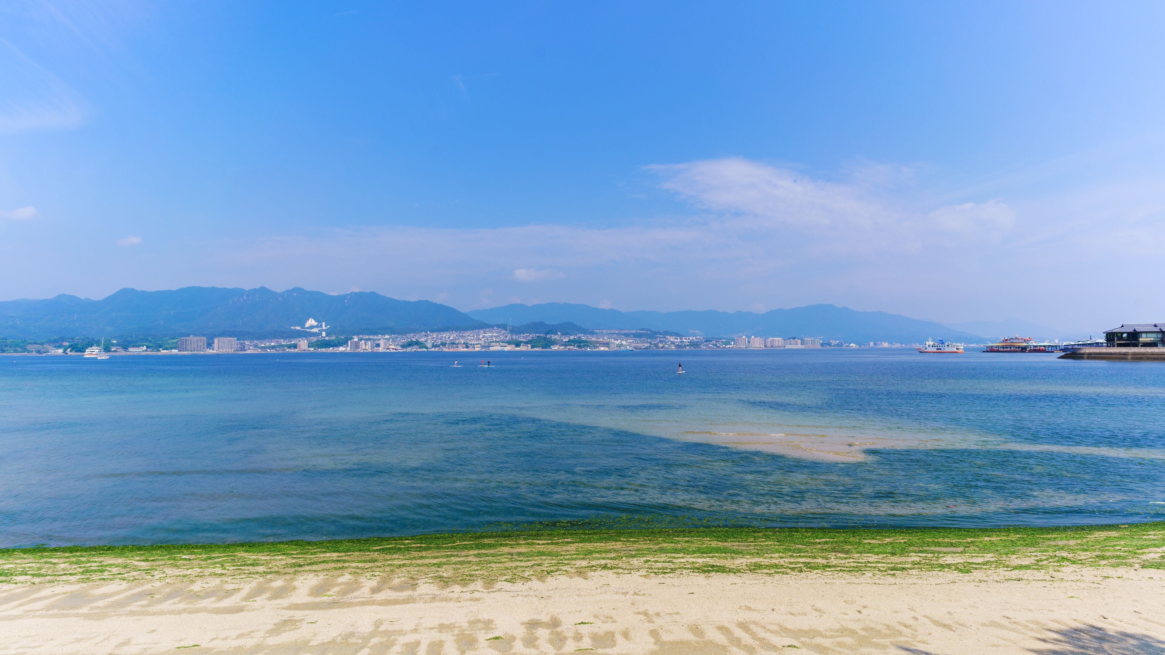 Paisagem costeira com céu claro, mar calmo e montanhas ao longe.