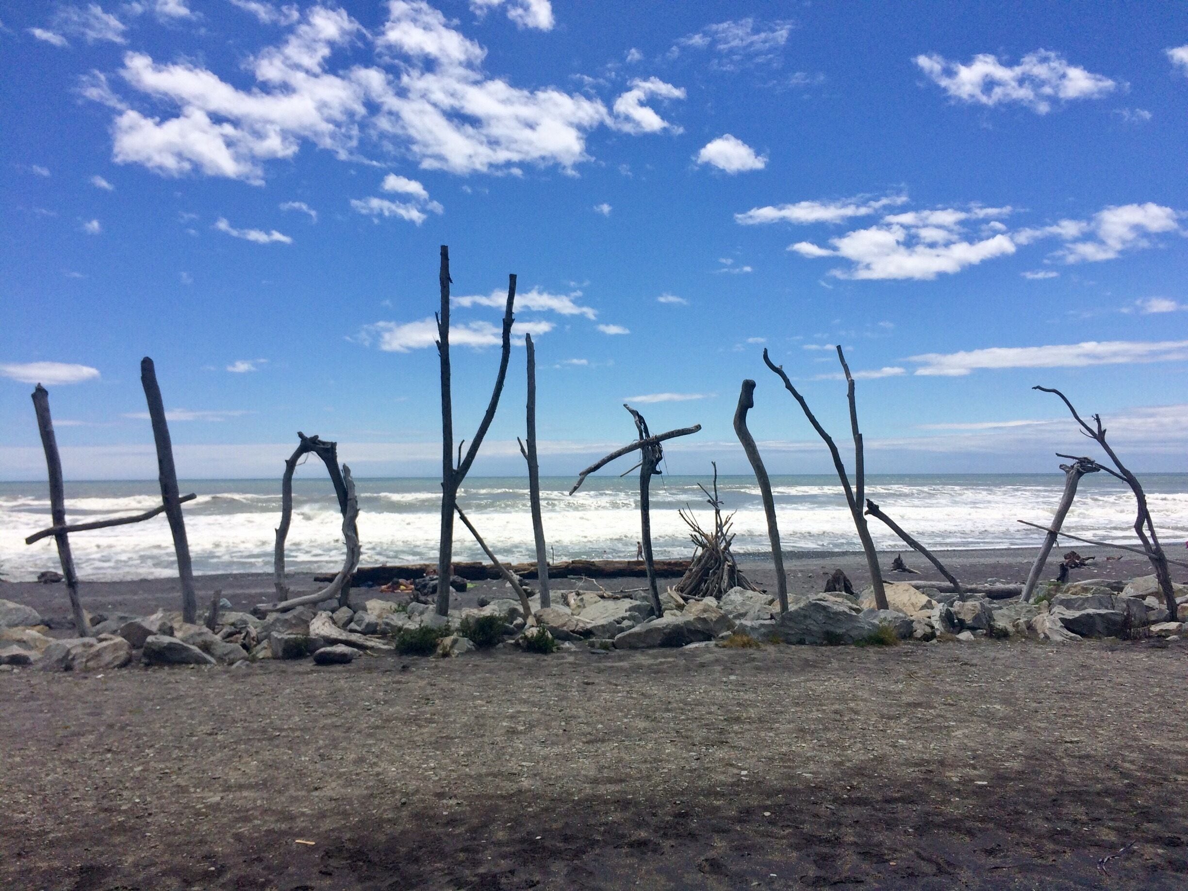 Praia com destroços de madeira e rochas sob um céu claro.