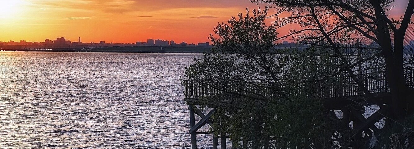 Um pôr do sol sobre a água, com um cais e árvores ao primeiro plano.