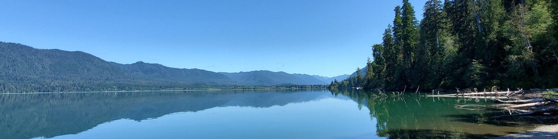 Um lago calmo com reflexos nítidos, rodeado por montanhas e uma floresta densa.