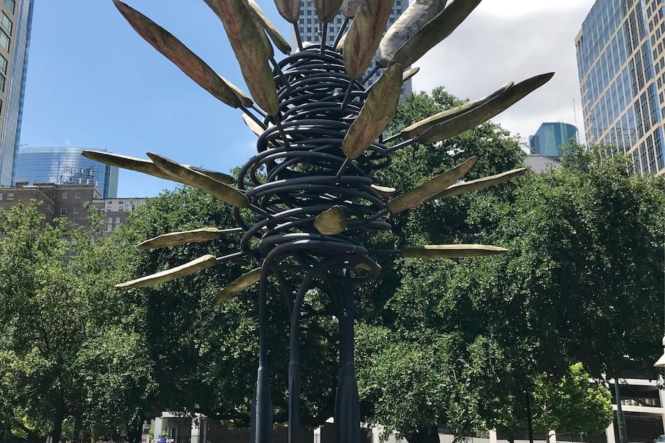 Escultura com elementos semelhantes a folhas em um parque urbano, com edifícios altos ao fundo.