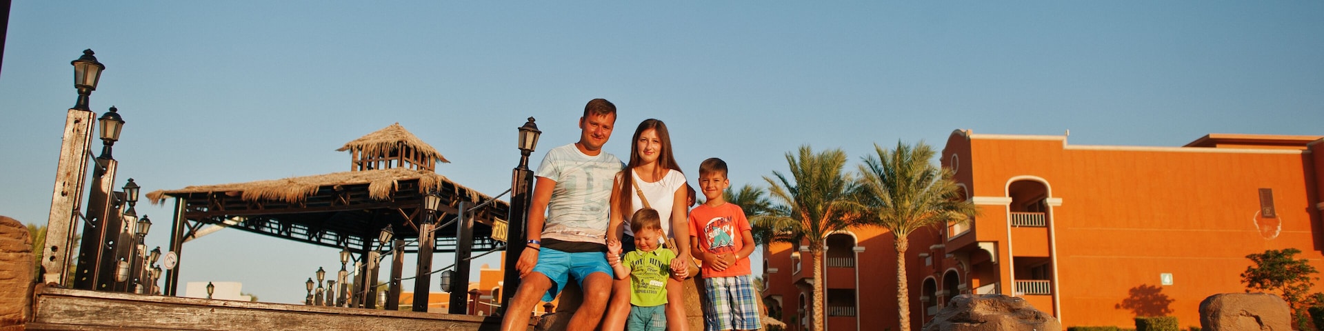 Uma família posando em frente a uma construção com telhado de palha, com um céu azul claro e edifícios alaranjados ao fundo.