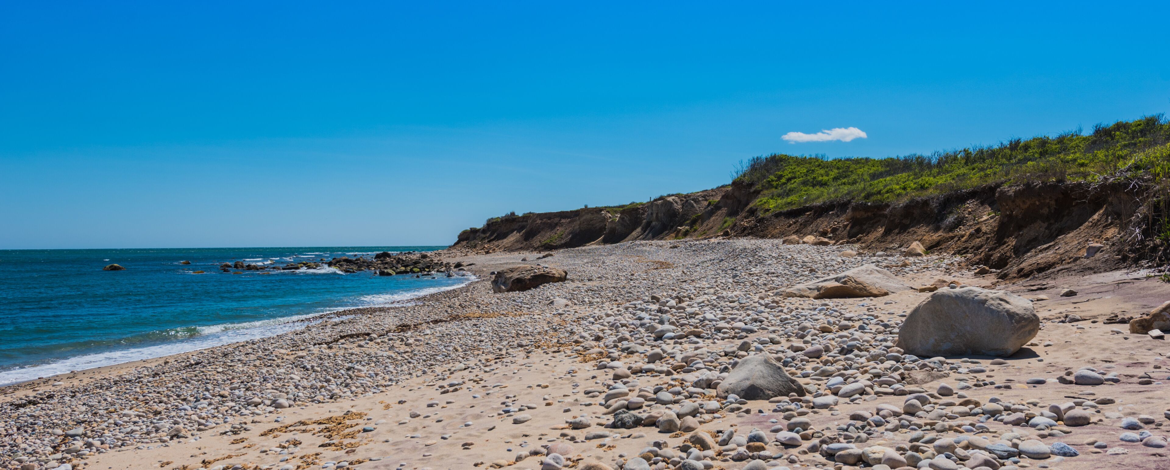 Uma praia de pedras com grandes rochas, céu azul límpido e um penhasco com vegetação.