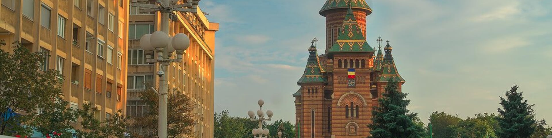 Um edifício histórico com uma cúpula verde e um campanário, rodeado por um jardim bem cuidado e edifícios modernos.