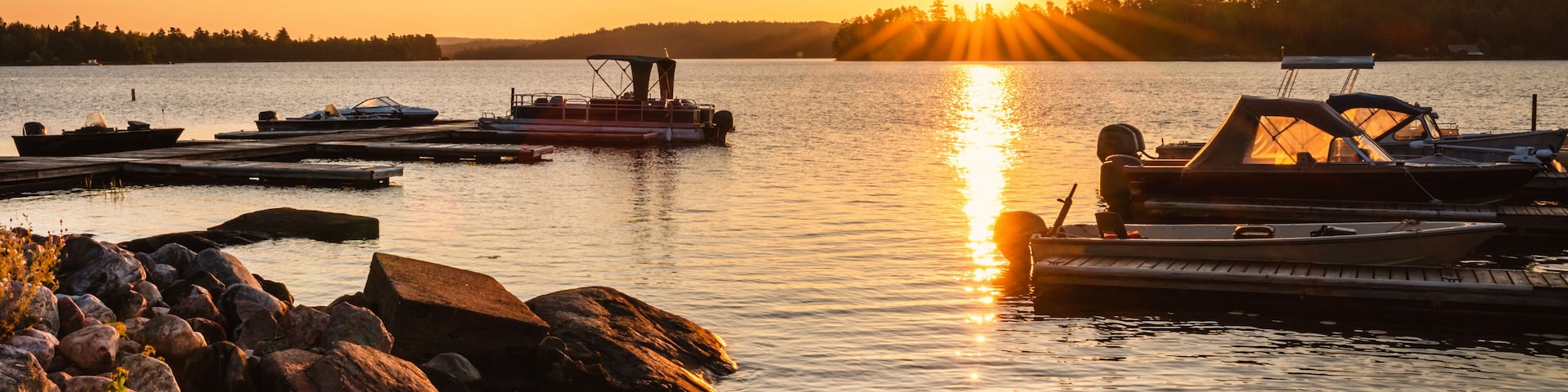 Um pôr do sol sobre um lago calmo, com barcos atracados, cercado por rochas e árvores.