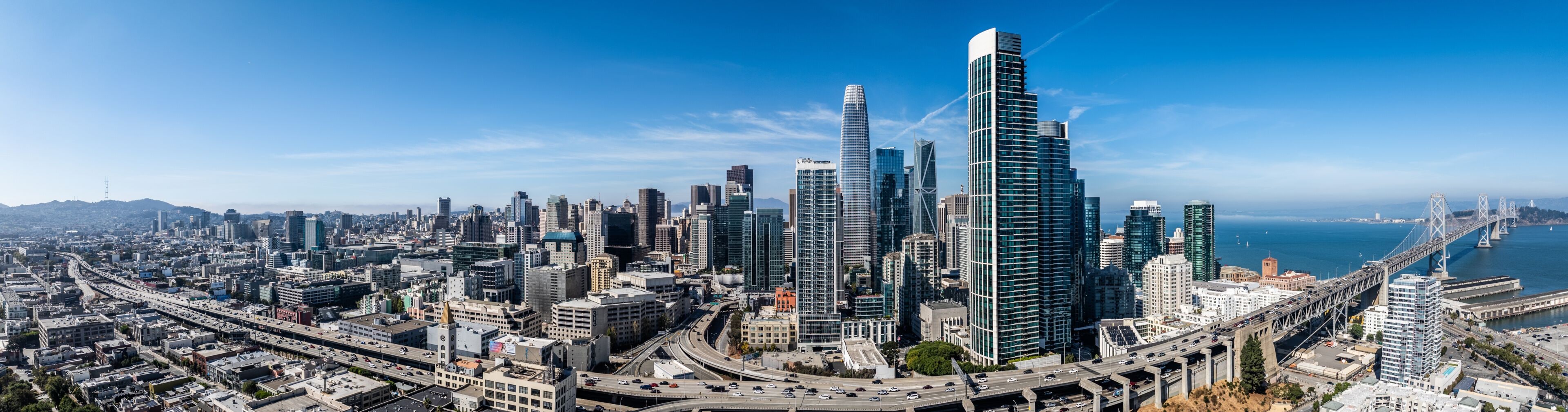 Paisagem urbana com um arranha-céu imponente, edifícios densos e uma ponte sobre a água.