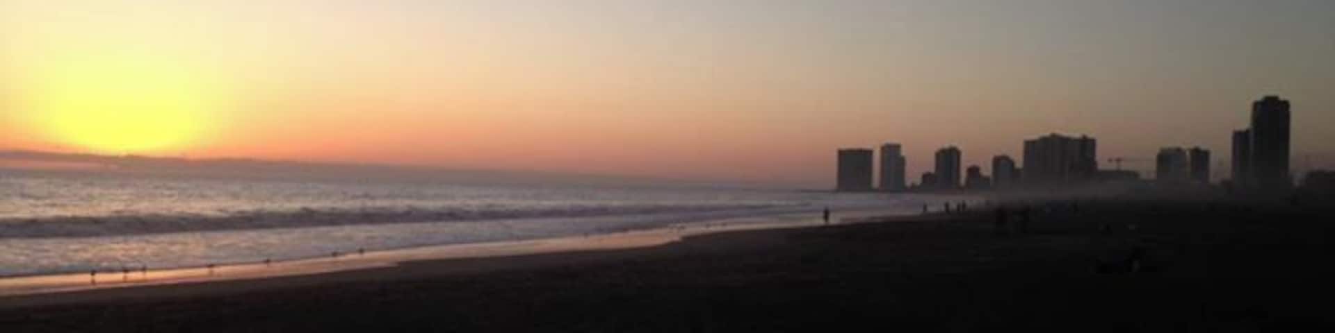 Uma praia ao pôr do sol com silhuetas de edifícios ao fundo.