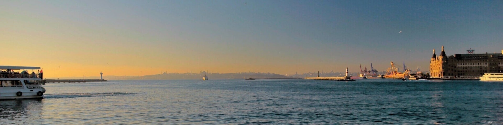 Um pôr do sol sobre um rio calmo, com um barco, o horizonte de uma cidade ao longe e um edifício imponente com torres.