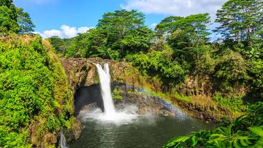 Uma cascata deságua num rio, rodeado por vegetação exuberante e floresta densa.
