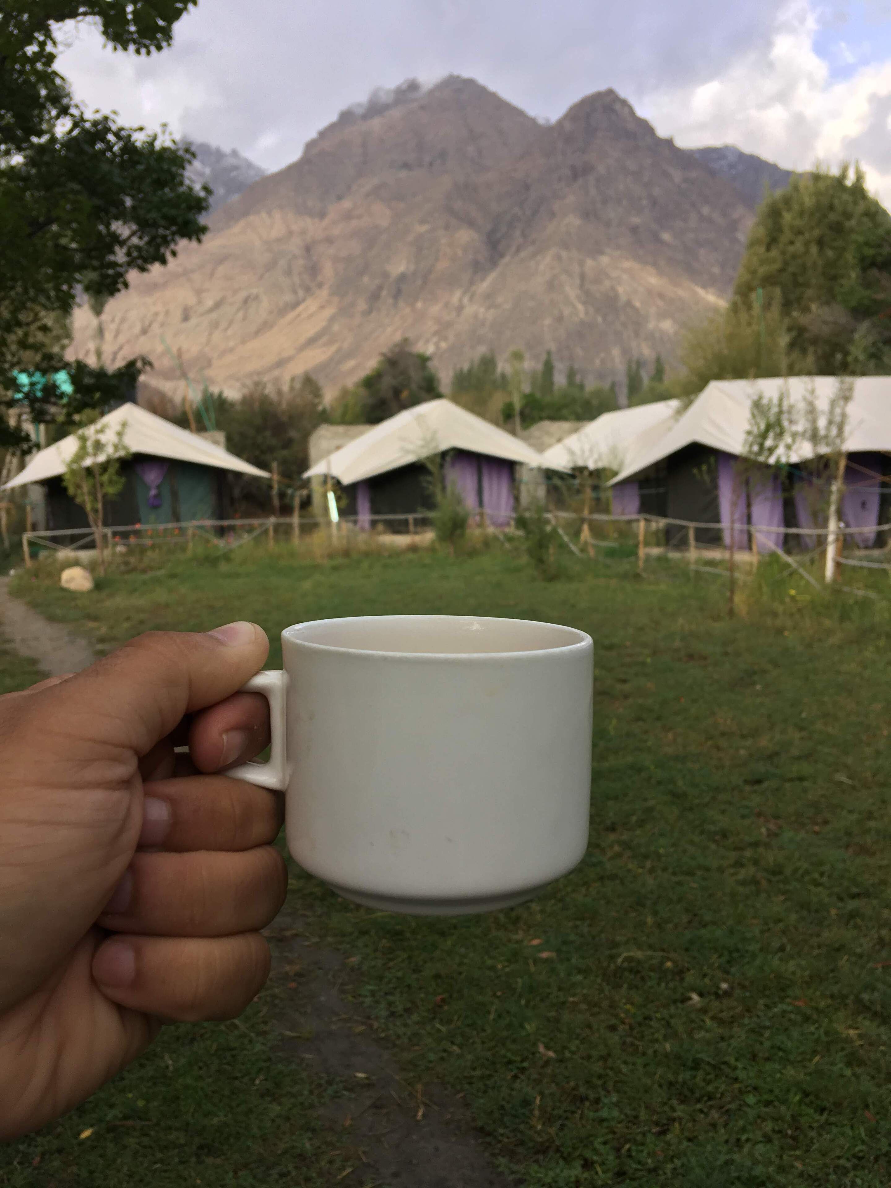 Mão segurando uma caneca branca diante de uma paisagem montanhosa com várias yurtas.
