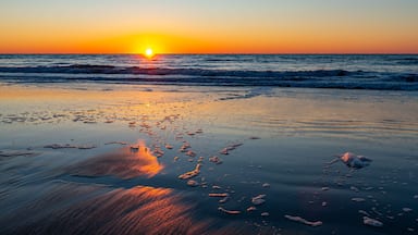 Um pôr do sol sobre uma praia com ondas e pegadas na areia.