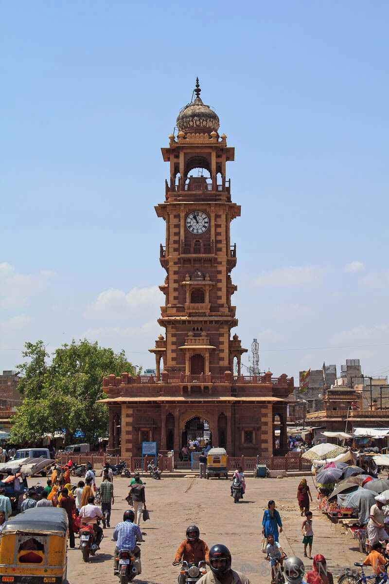 Uma torre do relógio histórica com uma cúpula, cercada por uma área de mercado movimentada com pessoas e veículos.