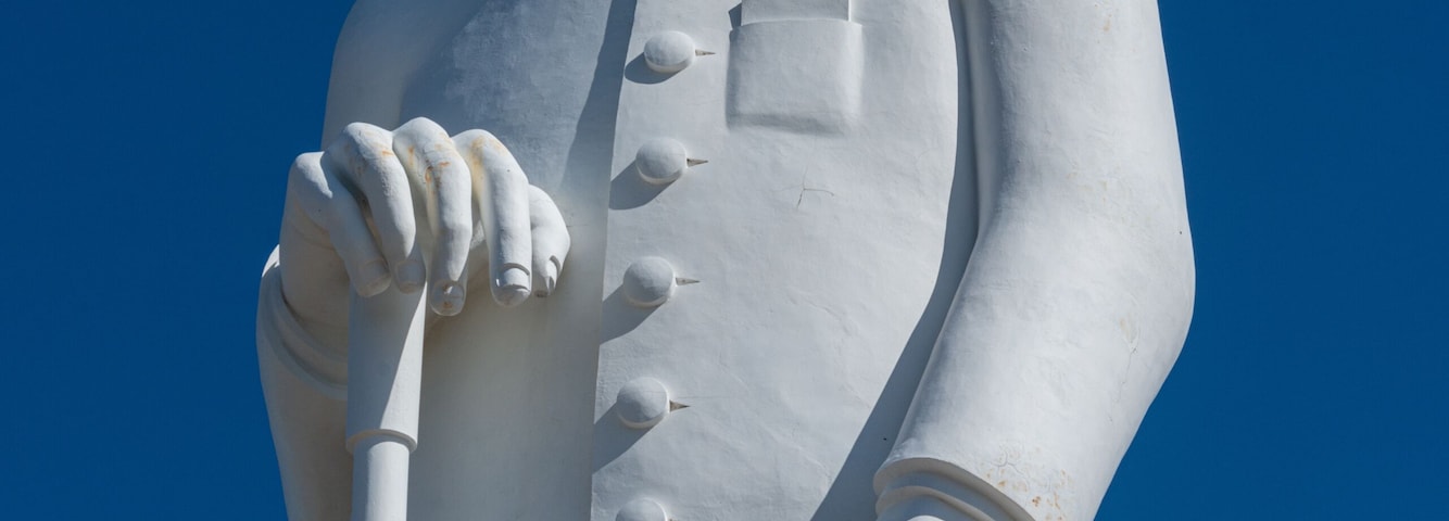 Uma estátua branca de um homem segurando um chapéu e uma bengala contra um céu azul claro.