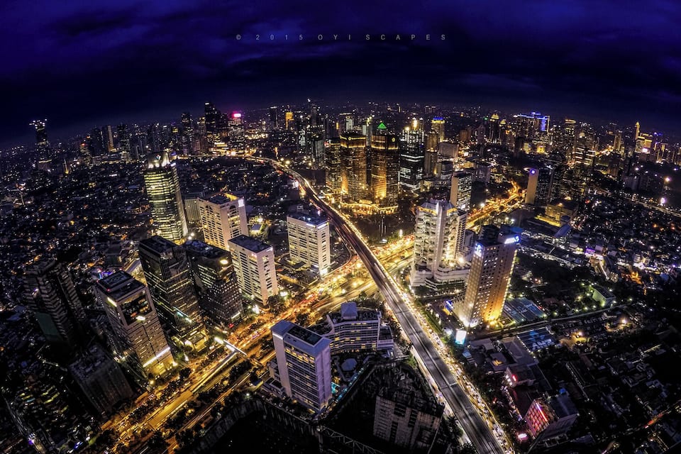 Vista aérea noturna de uma cidade densamente povoada, com arranha-céus iluminados e rodovias movimentadas.