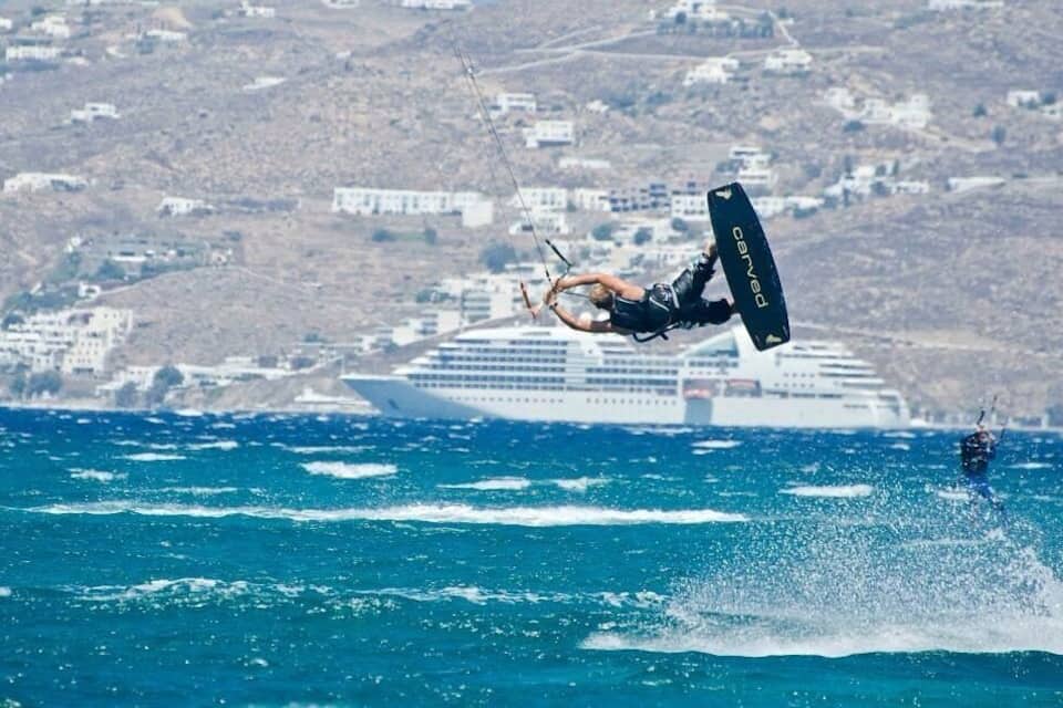 Uma pessoa praticando kitesurf sobre a água, com um cruzeiro ao fundo.