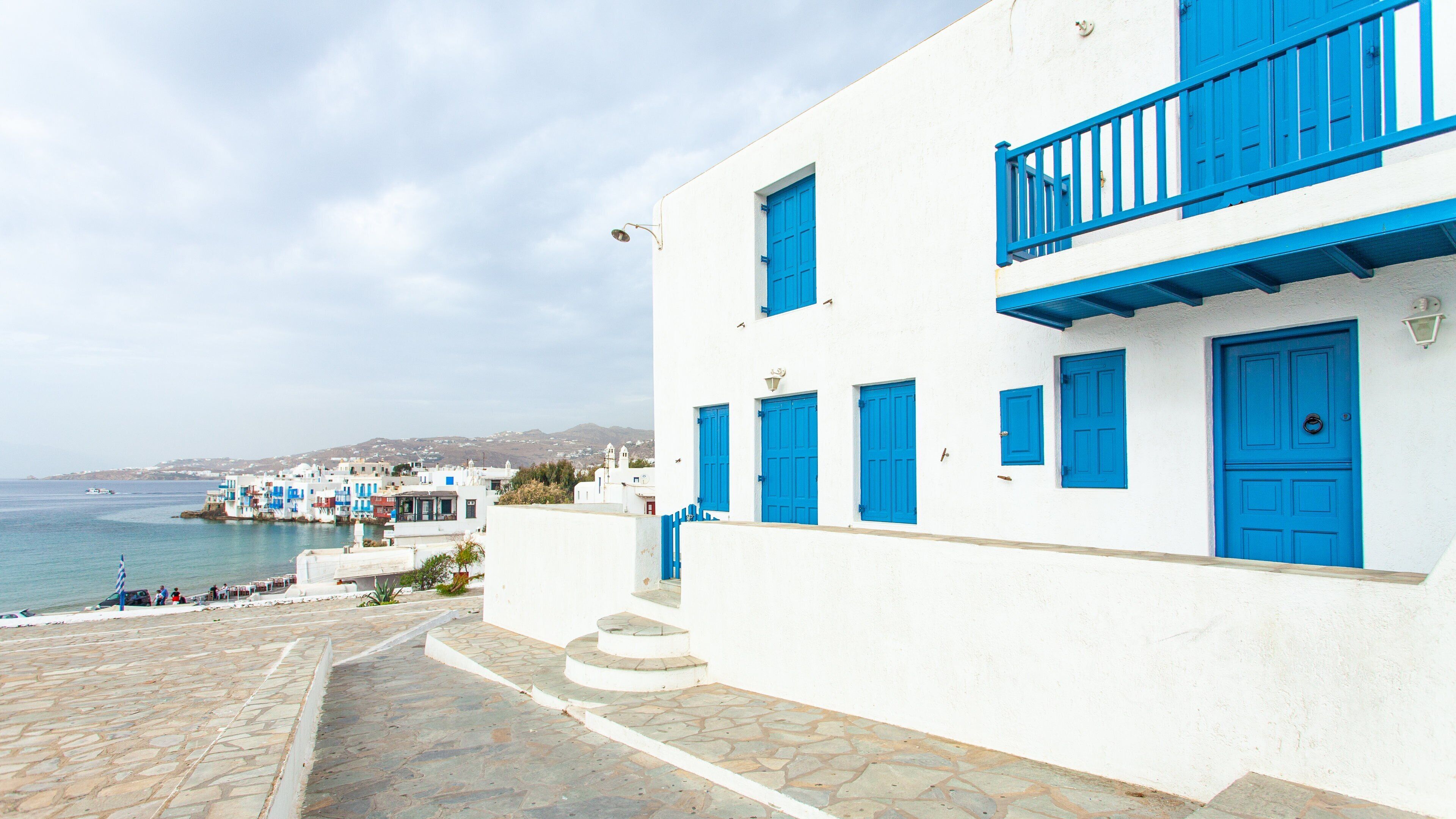 Um edifício branco com portas e janelas azuis, uma escadaria de pedra e vista para o mar e colinas ao longe.