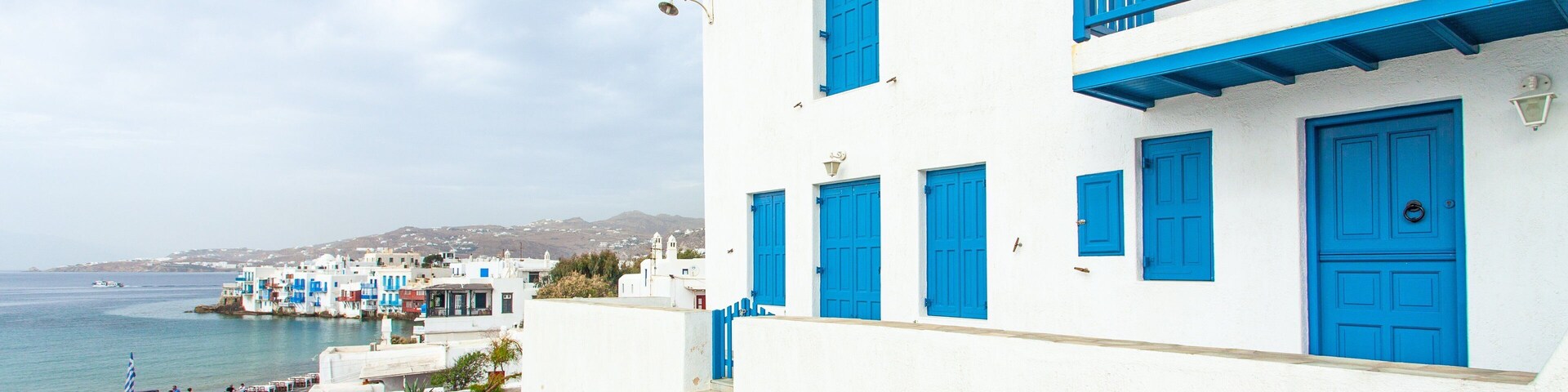 Um edifício branco com portas e janelas azuis, uma escadaria de pedra e vista para o mar e colinas ao longe.