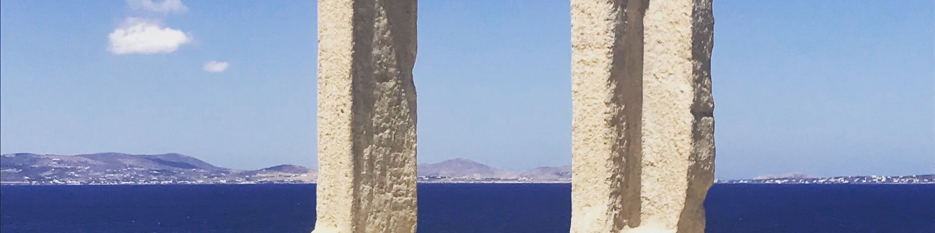 Ruínas antigas com colunas, ao fundo o mar e montanhas distantes.