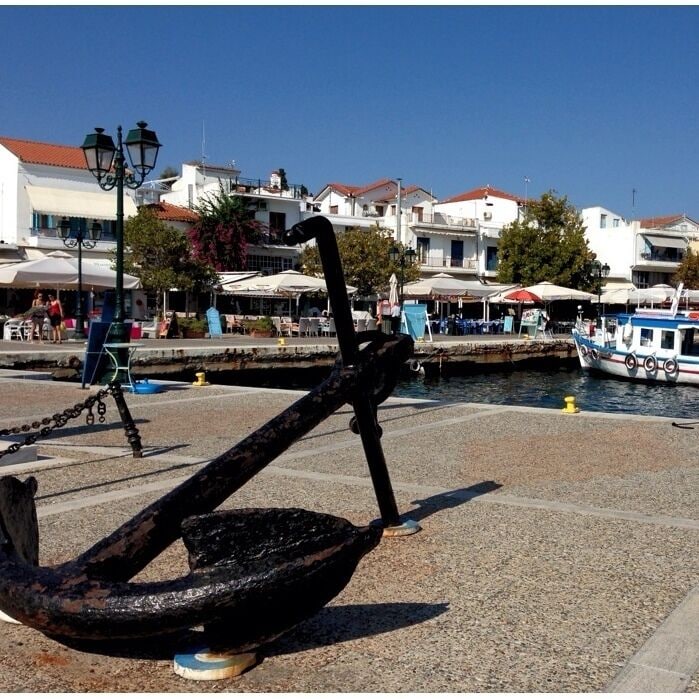 Margem com uma grande escultura de âncora, edifícios com telhados vermelhos e um barco azul atracado nas proximidades.