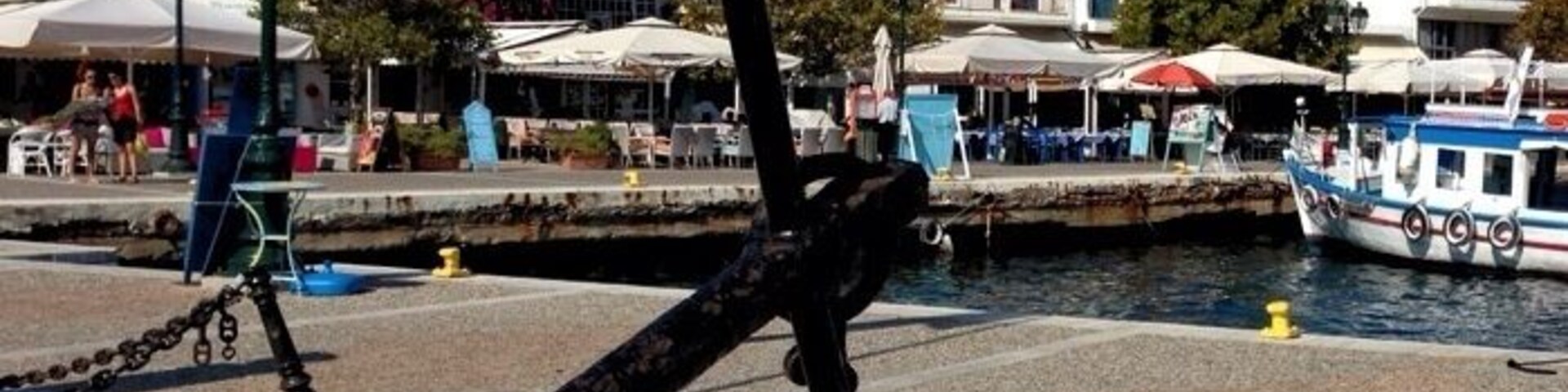 Margem com uma grande escultura de âncora, edifícios com telhados vermelhos e um barco azul atracado nas proximidades.