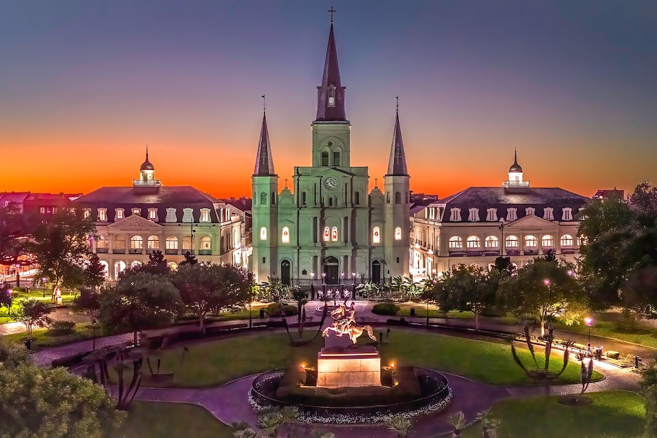 Uma catedral histórica com múltiplas torres, rodeada por jardins bem iluminados e uma fonte central.
