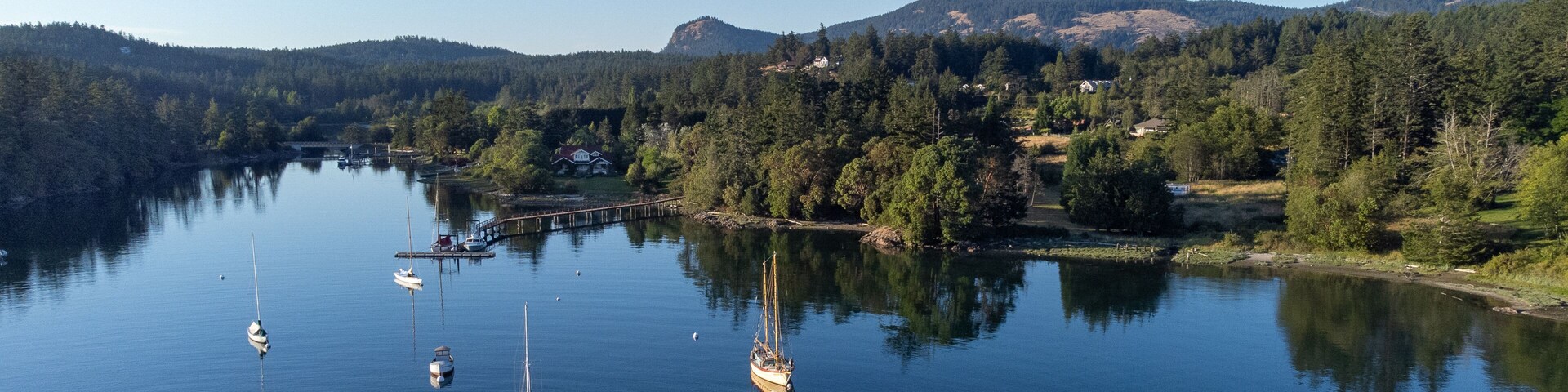 Um lago tranquilo com vários barcos à vela ancorados, rodeado por uma floresta densa e uma montanha ao fundo.