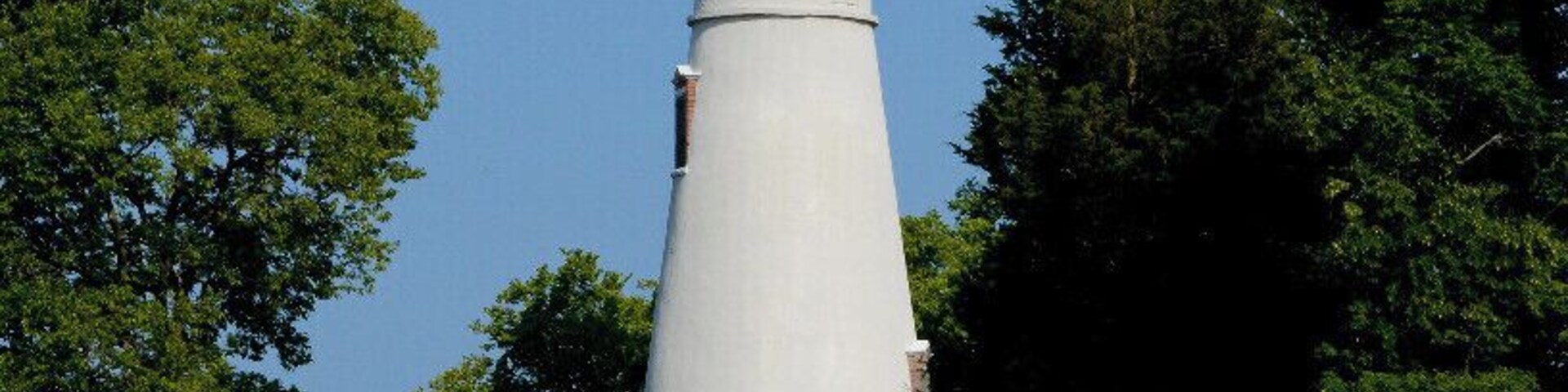 Um farol branco com a parte superior vermelha, rodeado por árvores e uma cerca branca de piquete.