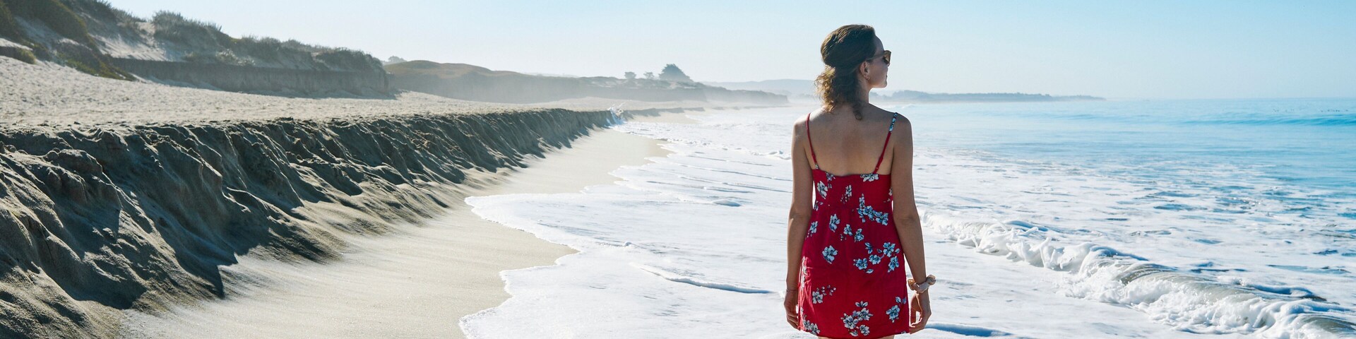 Uma mulher de vestido vermelho em uma praia arenosa, com ondas, céu claro e costa distante.