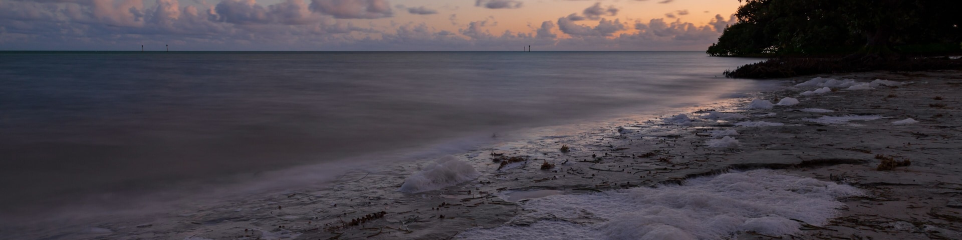 Uma praia com ondas espumosas, mar calmo e um céu de pôr do sol com nuvens.