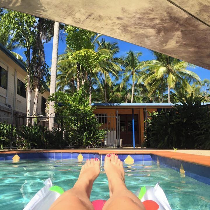 Uma piscina com uma pessoa relaxando em um flutuador, cercada por palmeiras e um edifício.
