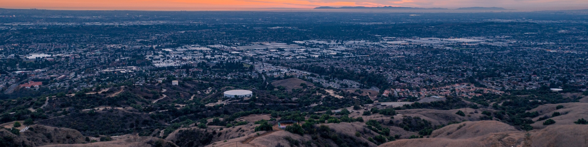Vista panorâmica de uma cidade ao pôr do sol, com colinas suaves em primeiro plano.